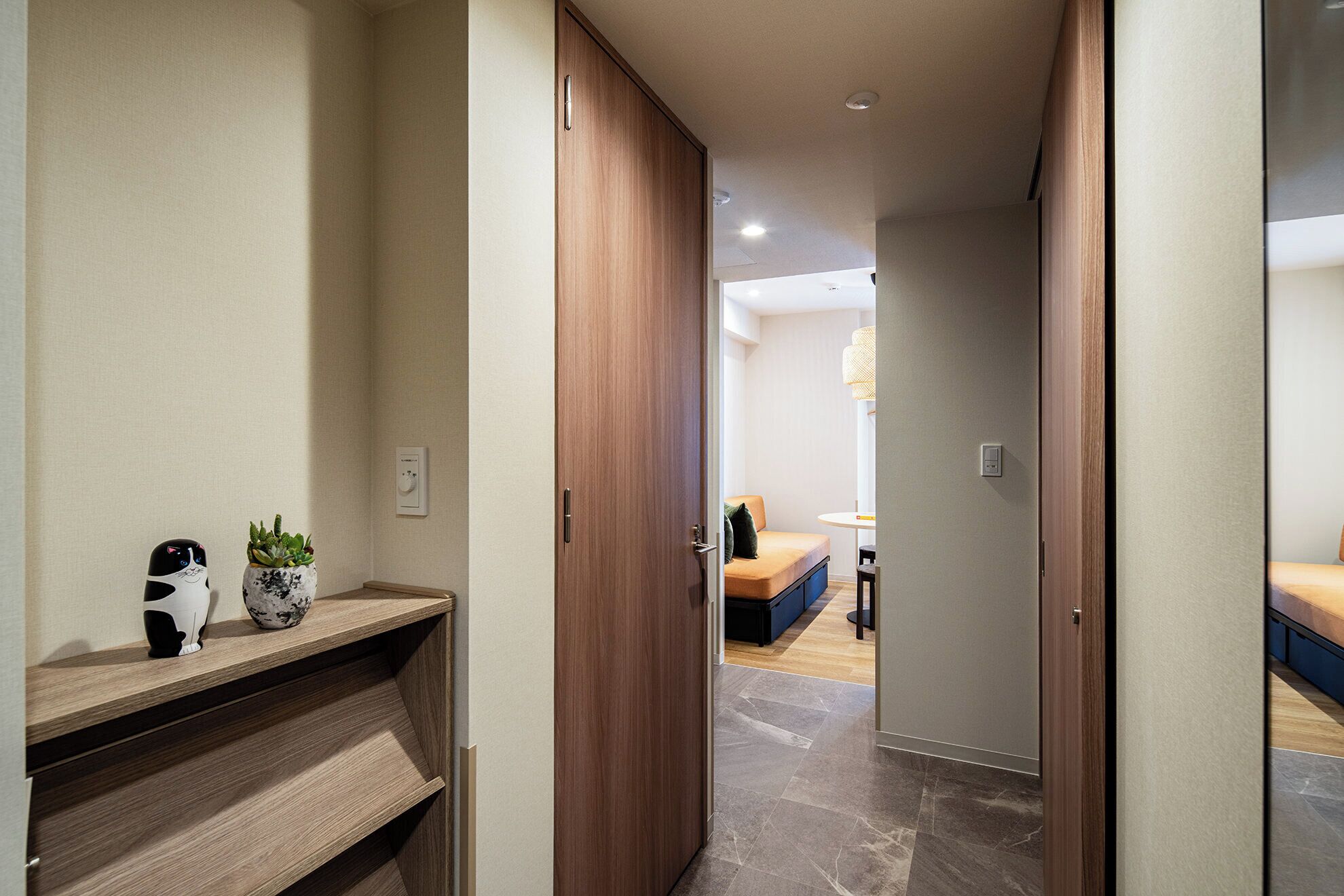 Um corredor com uma porta de madeira, uma prateleira com objetos decorativos e um vislumbre de um quarto com cama e mesa.