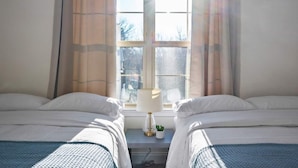 Quarto com duas camas, janela com cortinas, mesa de cabeceira com abajur e uma pequena planta.