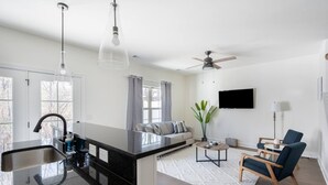 Uma cozinha moderna com bancada preta e pia de aço inoxidável, um sofá cinza com almofadas, uma mesa de centro e uma TV fixada na parede.
