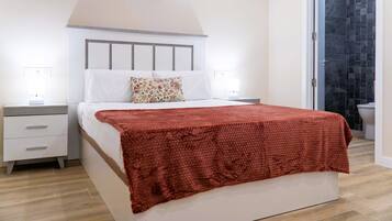 Quarto com cama, mesa de cabeceira e banheiro visível através de uma porta aberta.