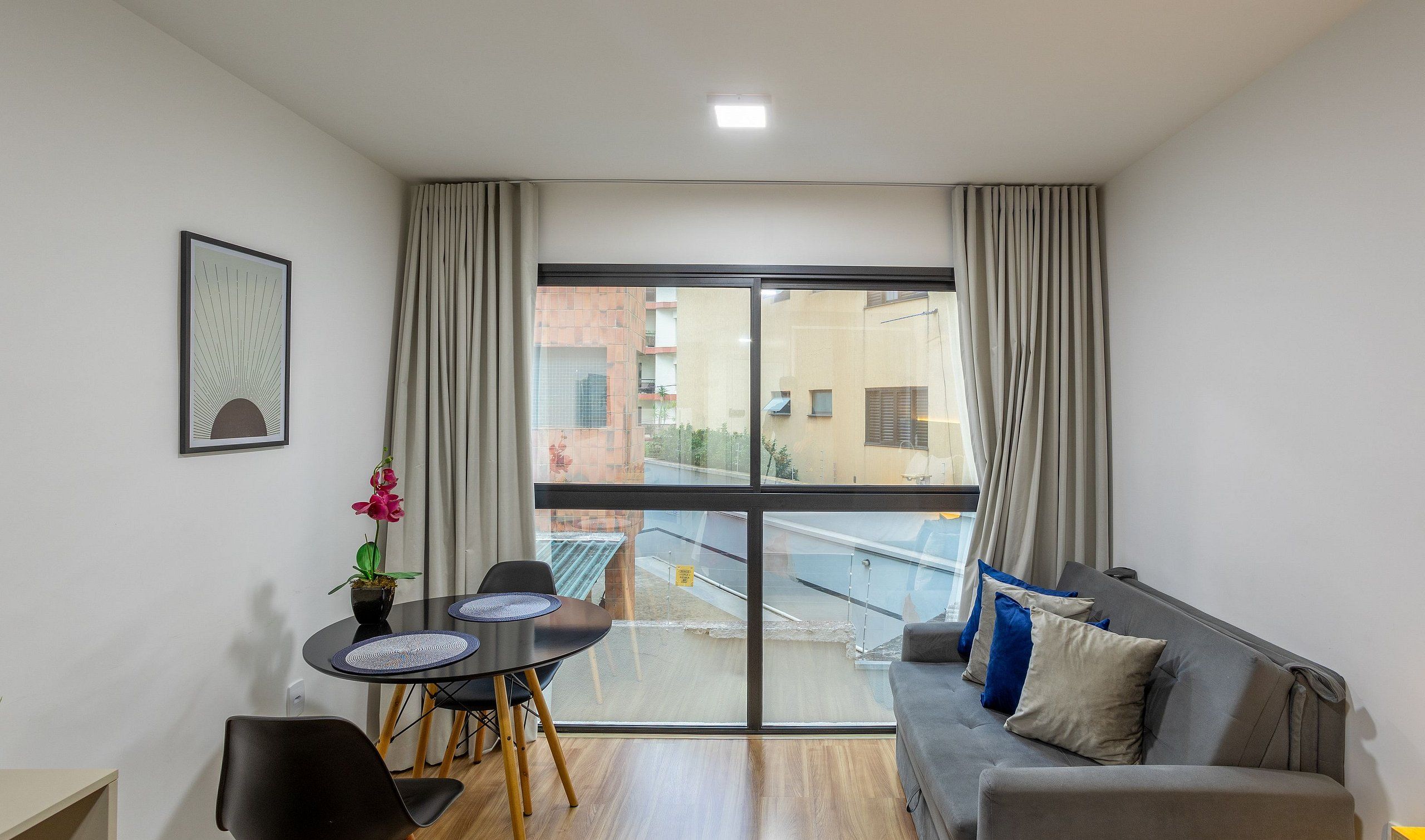 Uma sala de estar moderna com um sofá cinzento, uma mesa de jantar com cadeiras e uma janela grande com cortinas.