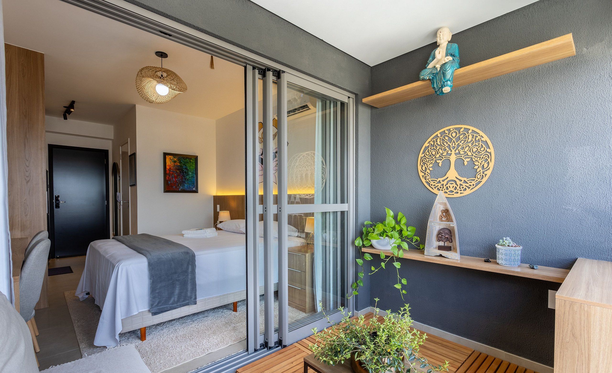 Quarto de hotel moderno com porta de vidro deslizante para uma varanda, cama com manta cinzenta, mesa pequena com planta e um objeto decorativo na parede.