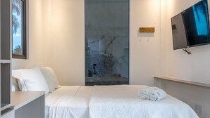 Quarto de hotel moderno com uma cama grande, uma televisão na parede e uma janela com vista para palmeiras.