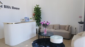 Uma área de recepção com um balcão branco, um sofá bege, uma mesa de centro redonda preta e uma planta em vaso com flores cor-de-rosa.