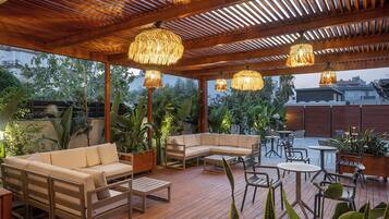 Um terraço ao ar livre com piso de madeira, um sofá, cadeiras e luminárias pendentes.