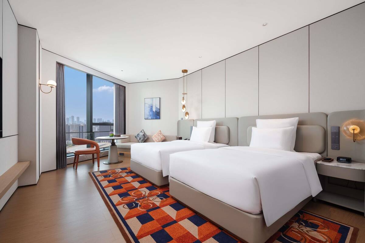Quarto de hotel moderno com duas camas, uma mesa de trabalho, uma cadeira e uma janela grande com vista para a cidade.