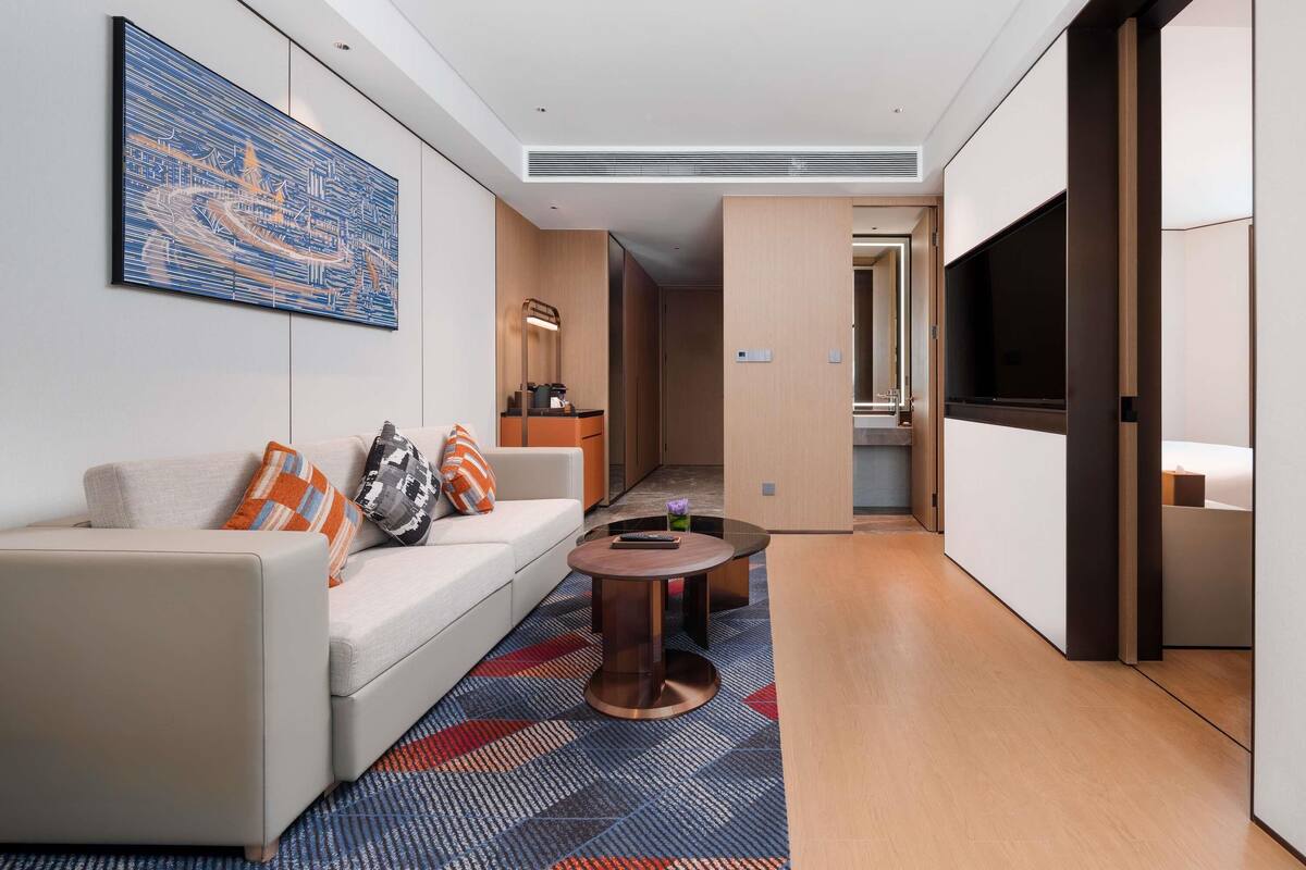 Quarto de hotel moderno com sofá, mesa de centro, pintura, espelho, televisão e banheiro visível por uma porta aberta.