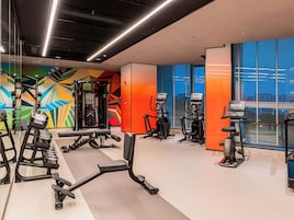 Uma academia com diversos equipamentos de exercício, uma parede colorida e janelas grandes.