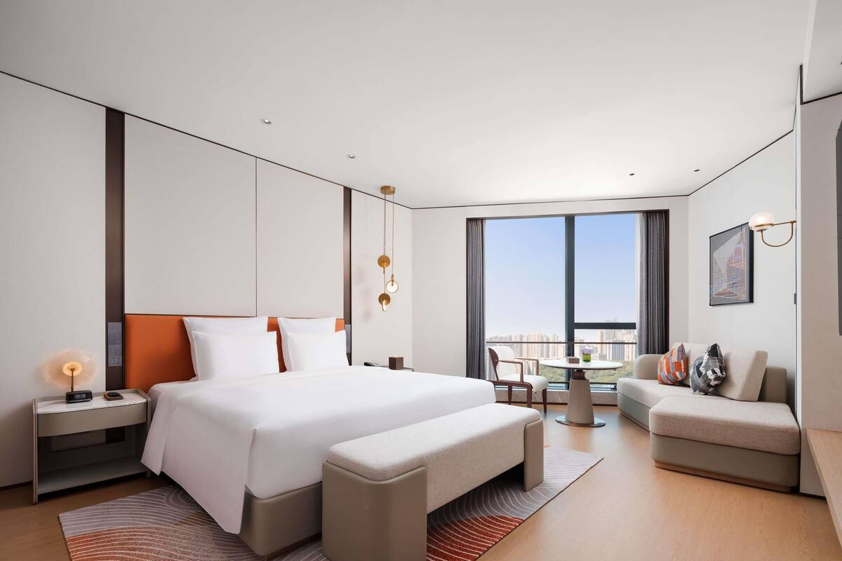 Quarto de hotel moderno com uma cama grande, um espaço de estar com mesa e cadeiras, e uma varanda com vista para a cidade.