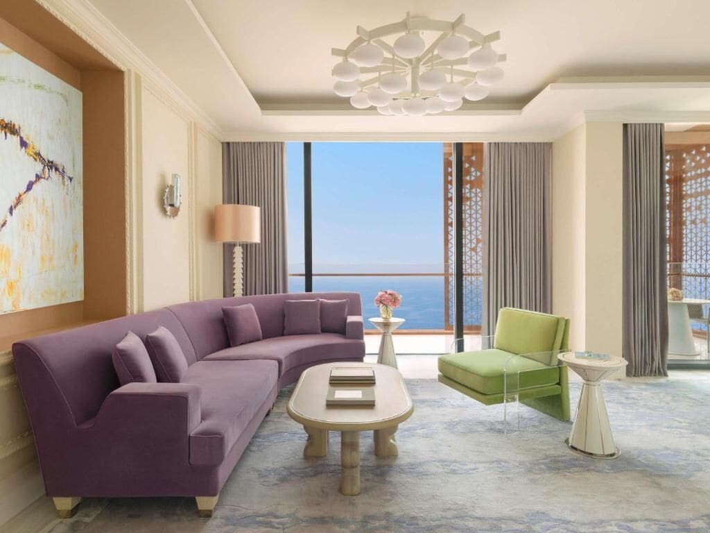 Quarto de hotel moderno com uma grande janela, um sofá roxo, uma poltrona verde, uma mesa de centro redonda e uma luminária decorativa no teto.