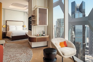 Quarto de hotel moderno com uma cama grande, área de estar com uma poltrona, uma mesa pequena e vista para a cidade através de amplas janelas.