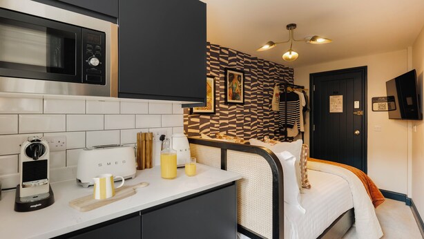 Cozinha compacta com micro-ondas, torradeira e cafeteira. Uma cama com cabeceira é visível ao fundo.