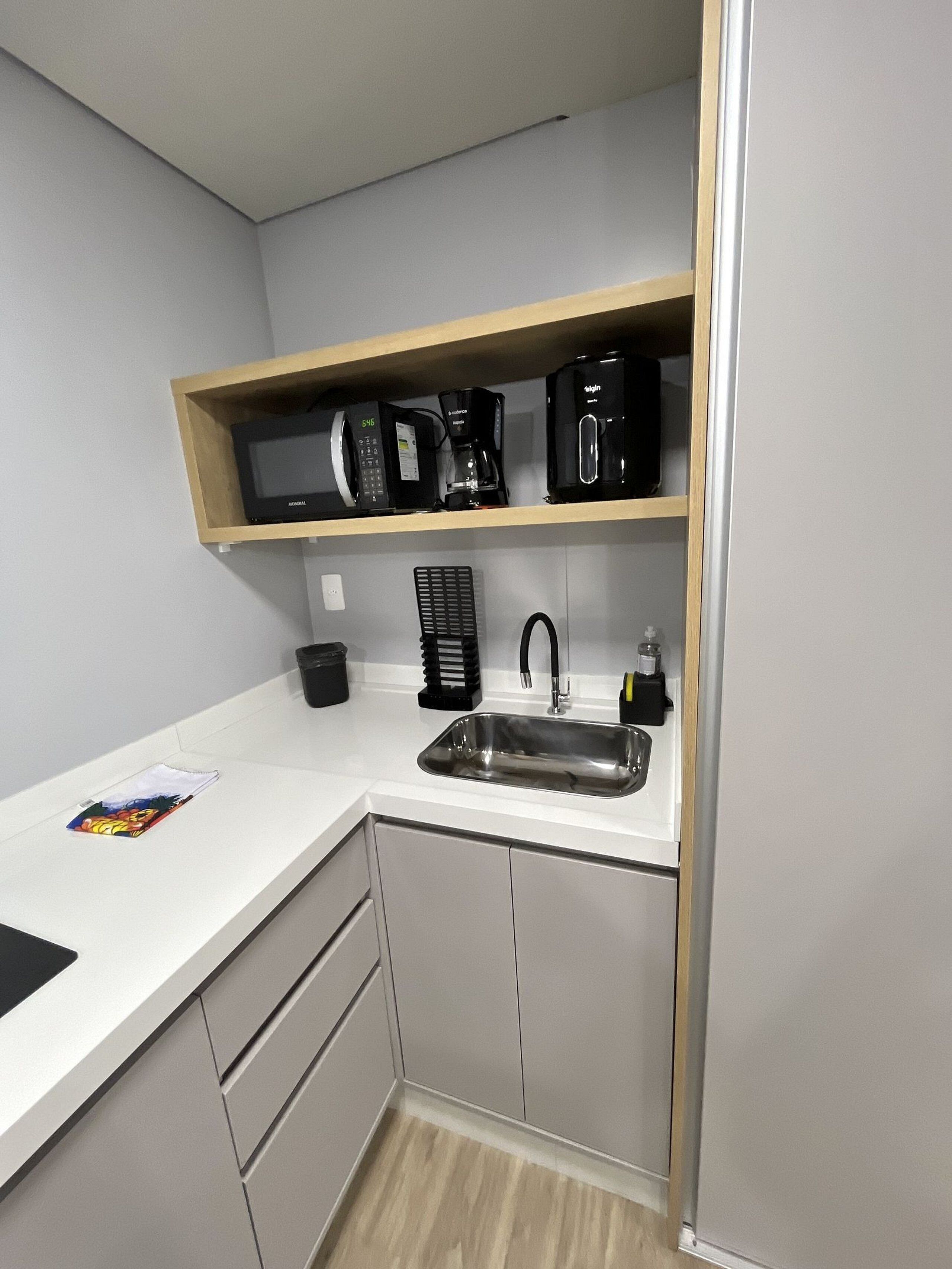Uma cozinha compacta com micro-ondas, cafeteira e chaleira em uma prateleira, pia com torneira e bancada com um único objeto.