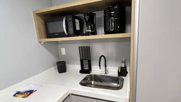 Uma cozinha compacta com micro-ondas, cafeteira e chaleira em uma prateleira, pia com torneira e bancada com um único objeto.