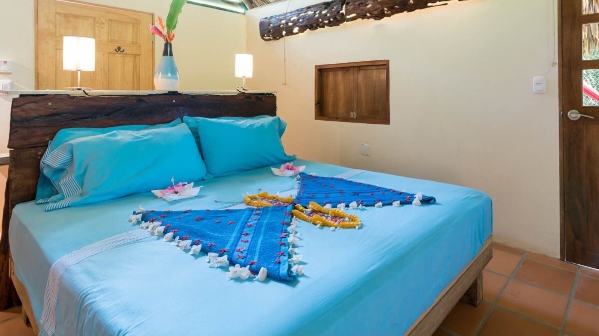 Uma cama bem arrumada com colcha e travesseiros azuis, cabeceira de madeira e uma pequena janela na parede.