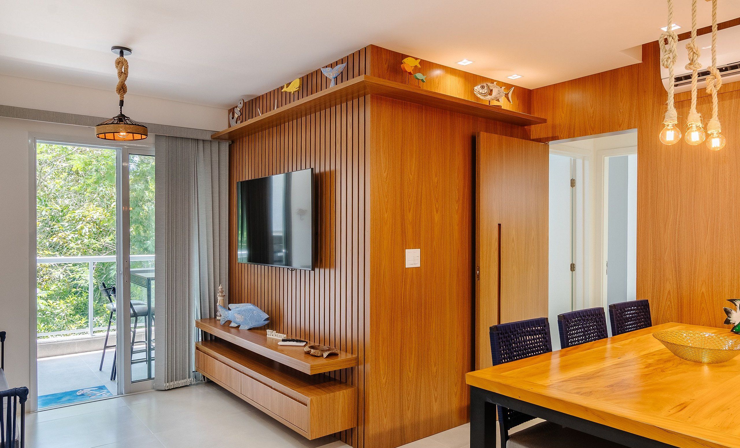 Uma sala de estar moderna com televisão de tela plana fixada em parede de madeira, área de jantar com mesa e cadeiras de madeira, e varanda com vista para o verde.