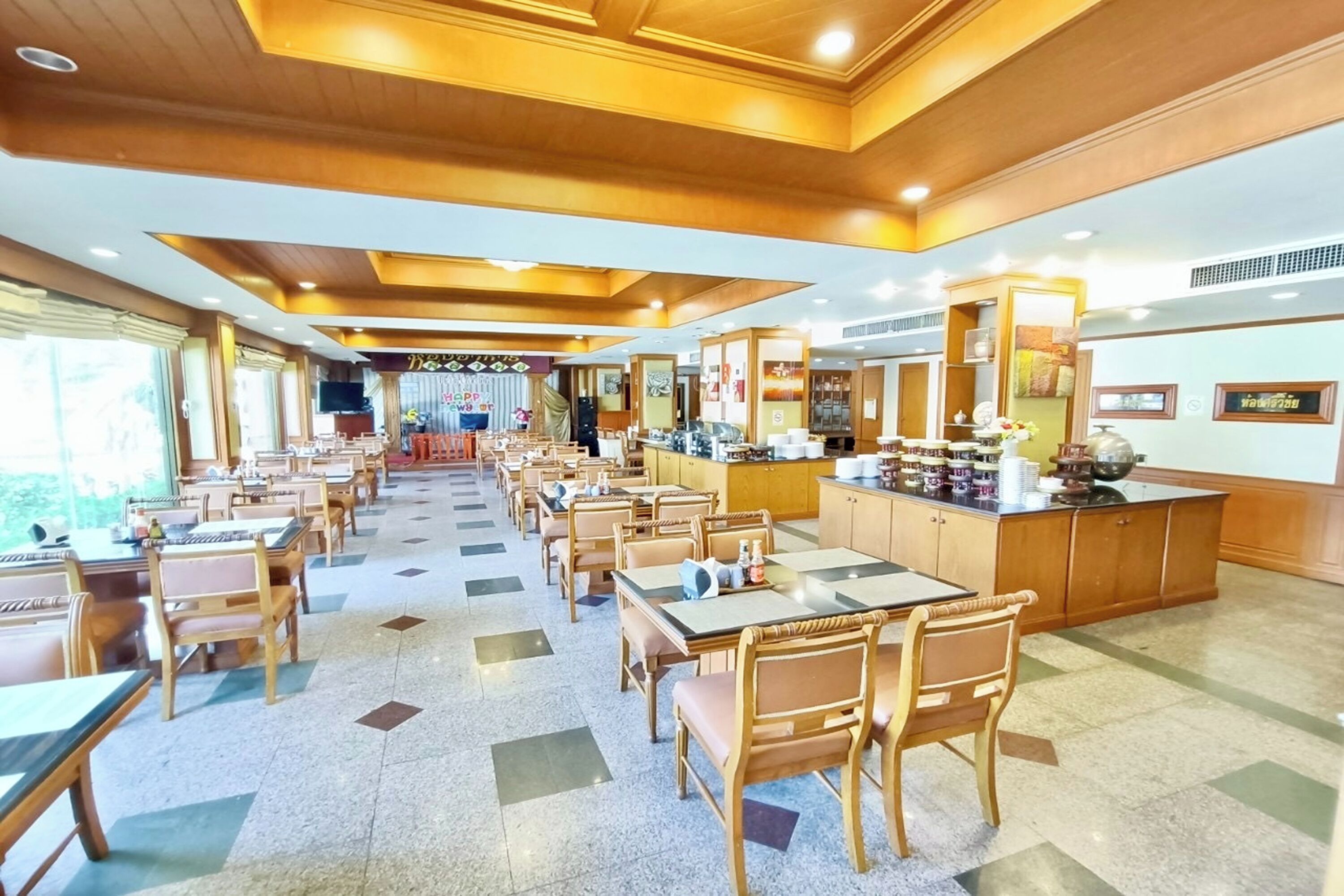 Ampla sala de jantar de hotel com várias mesas e cadeiras, um bar central e vista para a área externa.