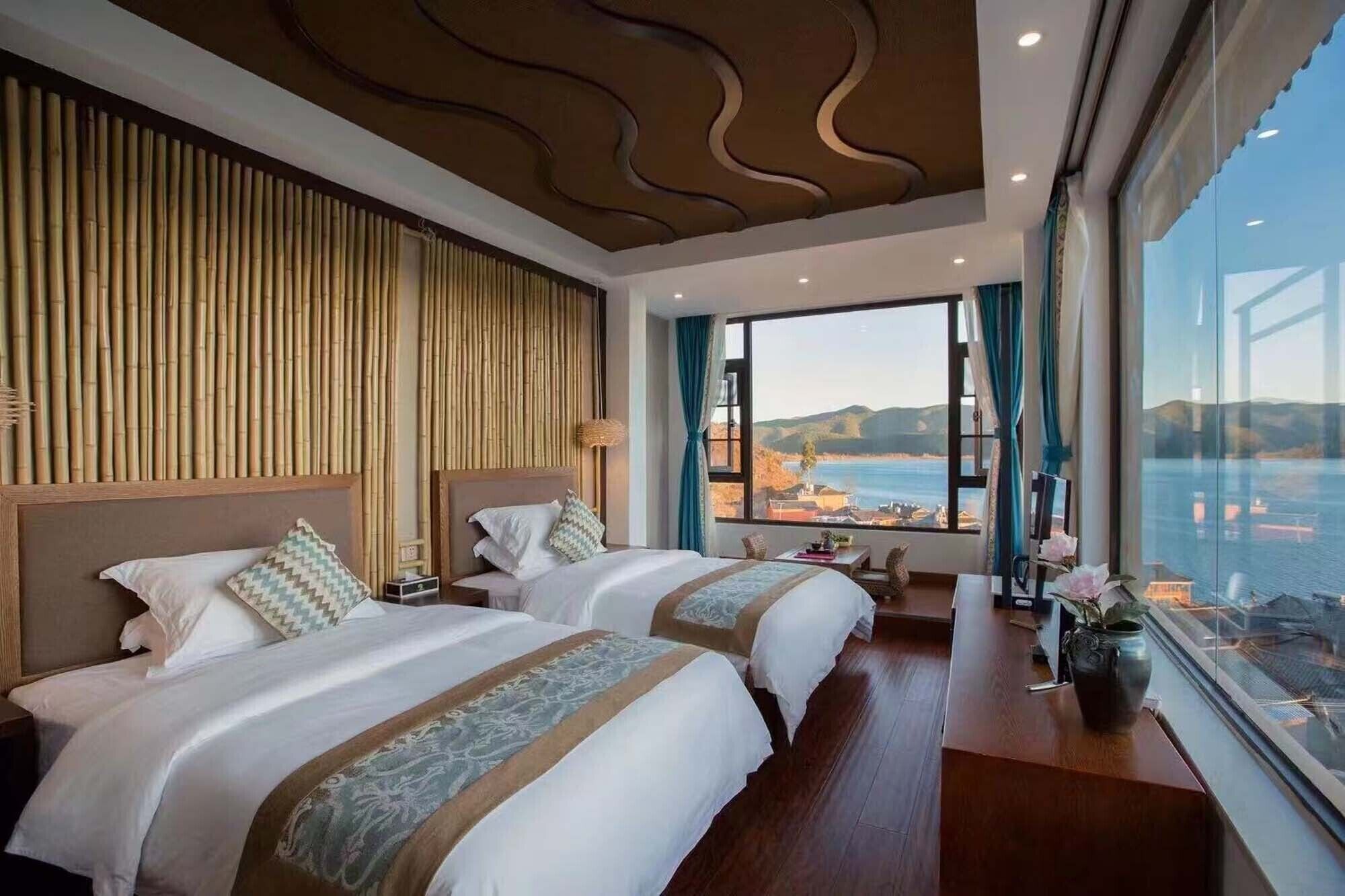 Quarto de hotel com duas camas, uma janela ampla com vista para um corpo d'água e colinas, e uma parede de bambu.