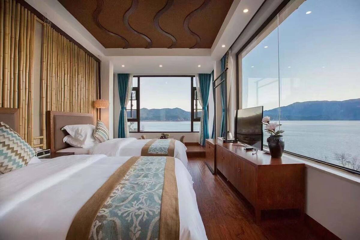 Quarto de hotel com duas camas, televisão e uma grande janela com vista para um lago e montanhas.