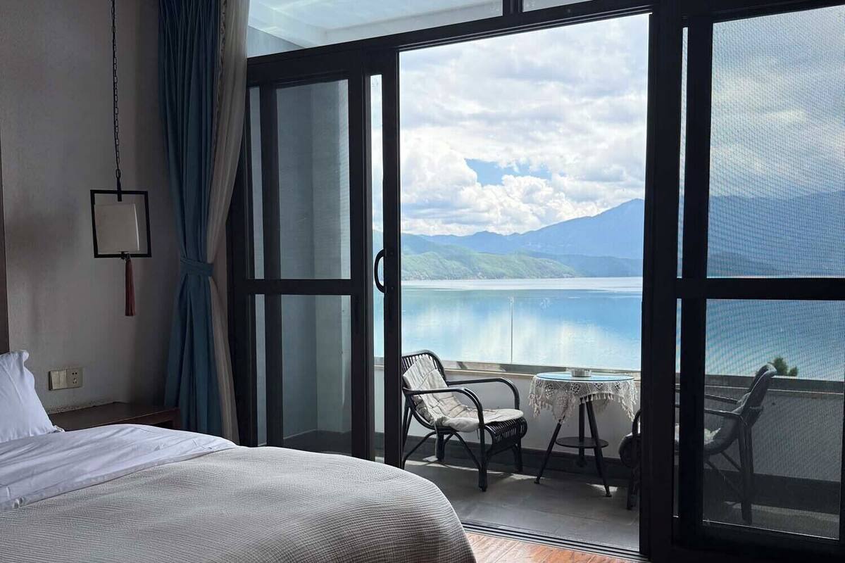 Quarto de hotel com cama, varanda com mesa e cadeiras, e vista para um lago e montanhas.