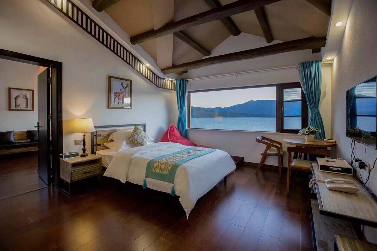 Quarto com cama, mesa de cabeceira, abajur e vista para um lago pela janela.