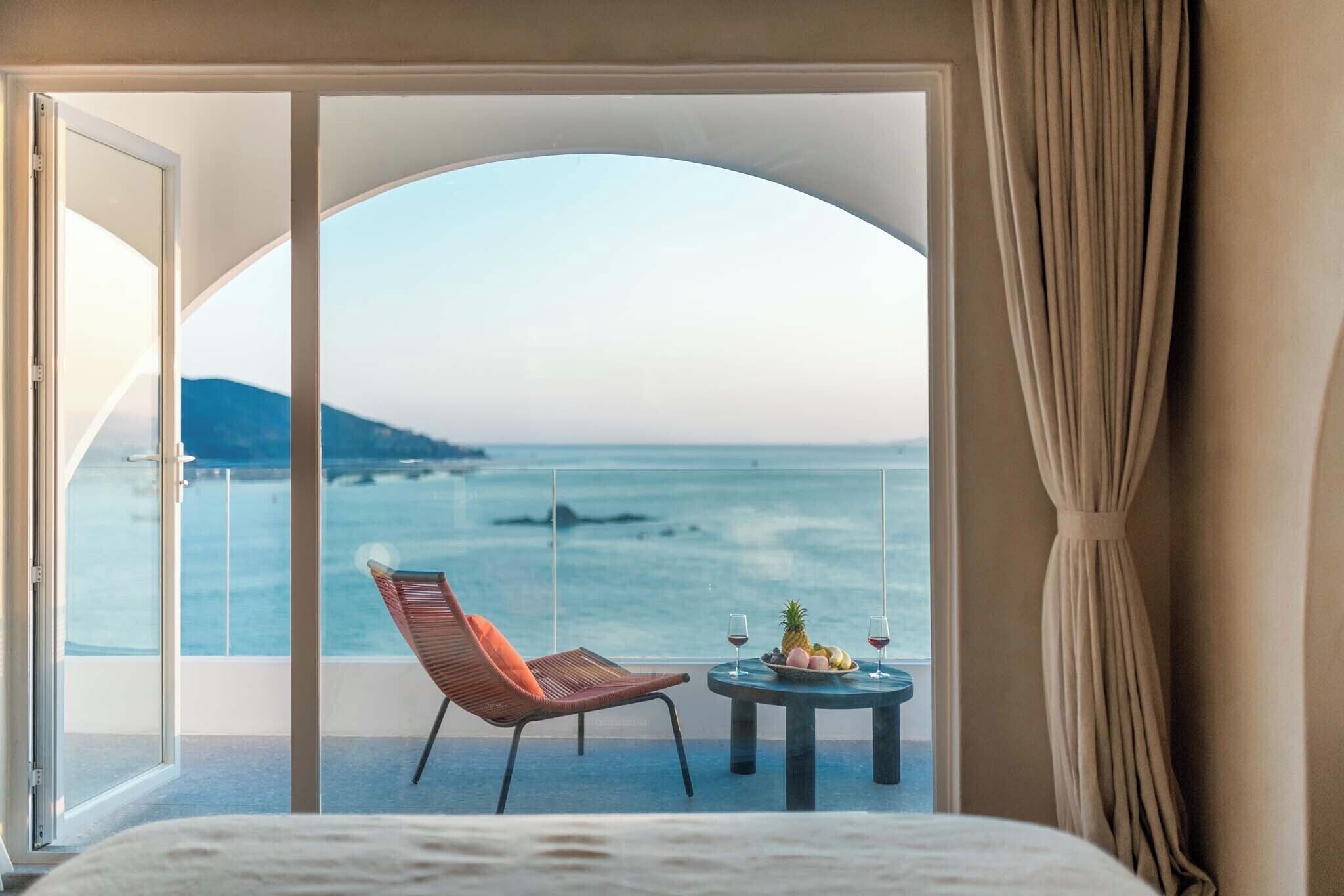 Varanda com vista para o mar, uma cadeira, uma mesa pequena com frutas e vinho, e uma cama.