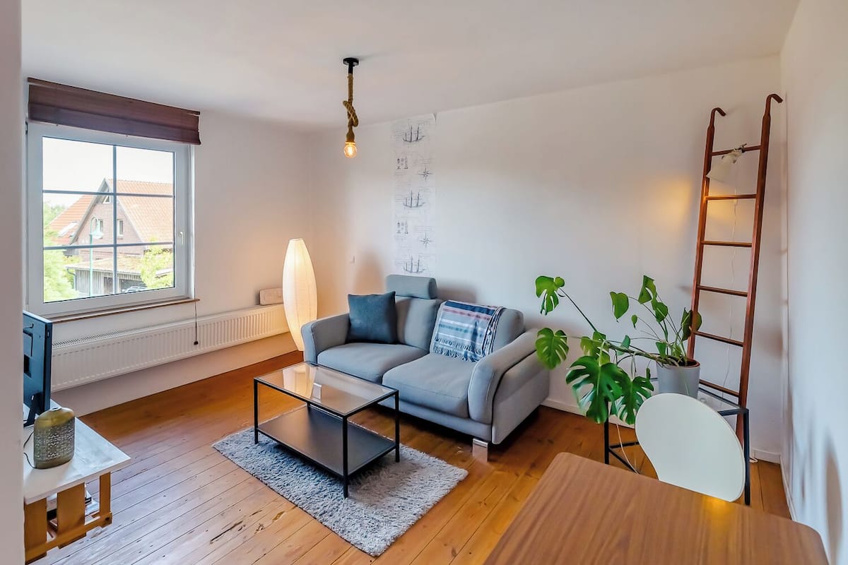 Uma sala de estar moderna com um sofá cinzento, uma mesa de centro de vidro, um abajur branco, uma escada de madeira e um vaso com planta.