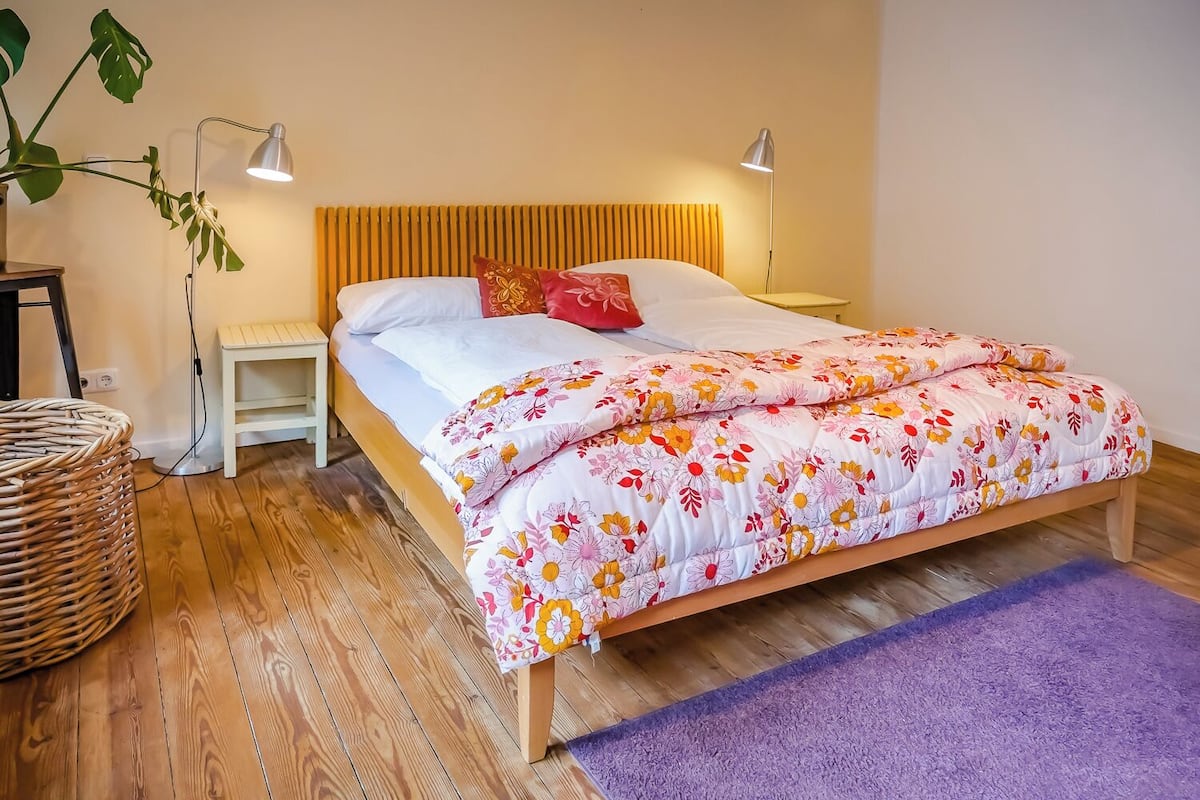 Uma cama bem arrumada com um edredom floral, uma mesa de cabeceira com abajur e uma planta em cesto de vime.