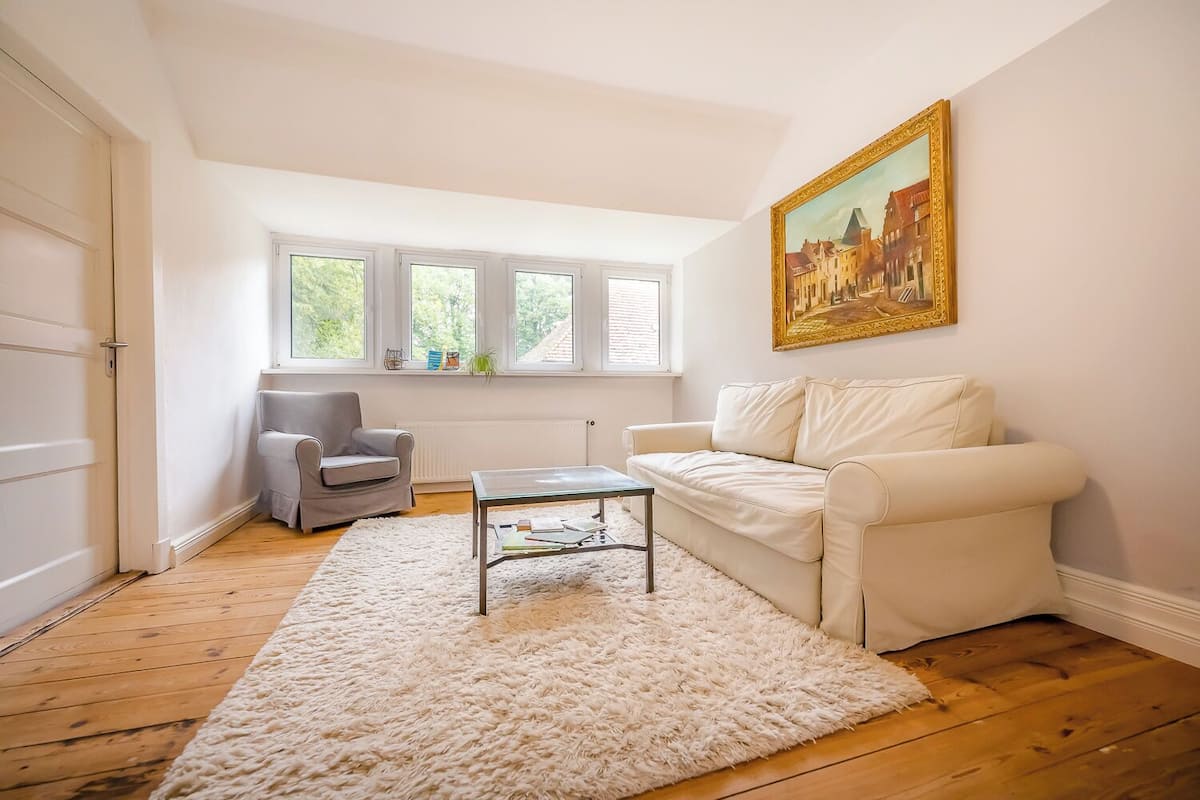 Sala de estar com sofá branco, poltrona cinza, mesa de centro de vidro e tapete fofo. Janelas com vista para vegetação.
