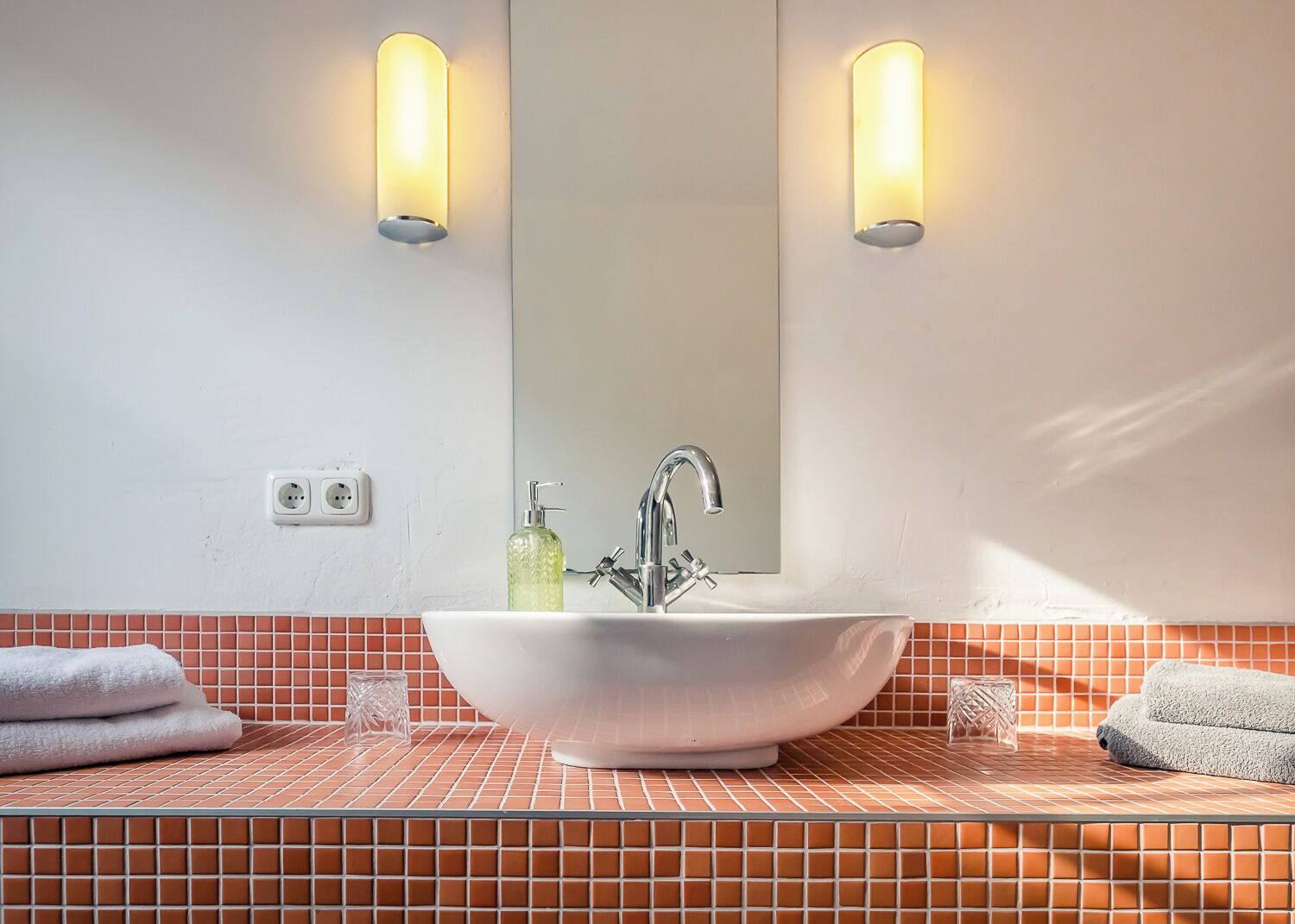 Casa de banho com uma pia branca, duas luzes na parede, um espelho e bancada revestida a azulejos.