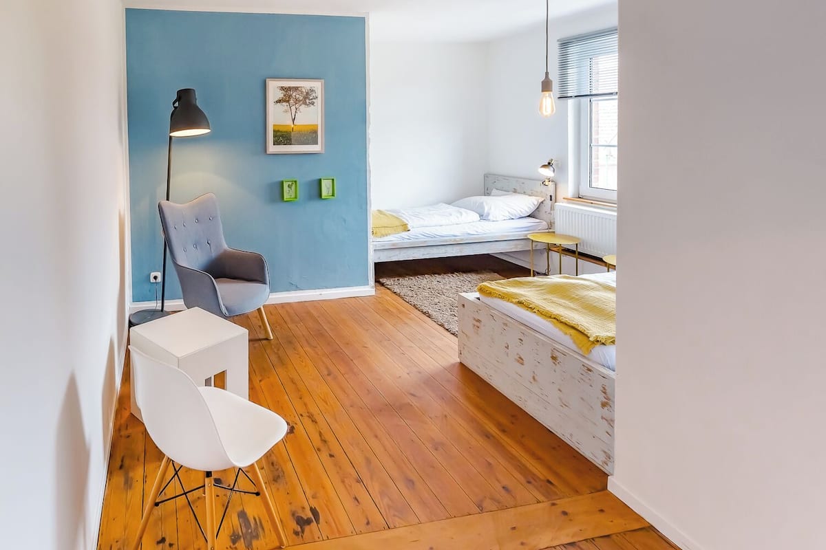 Quarto com cama, cadeira, escrivaninha, luminária e um quadro na parede.