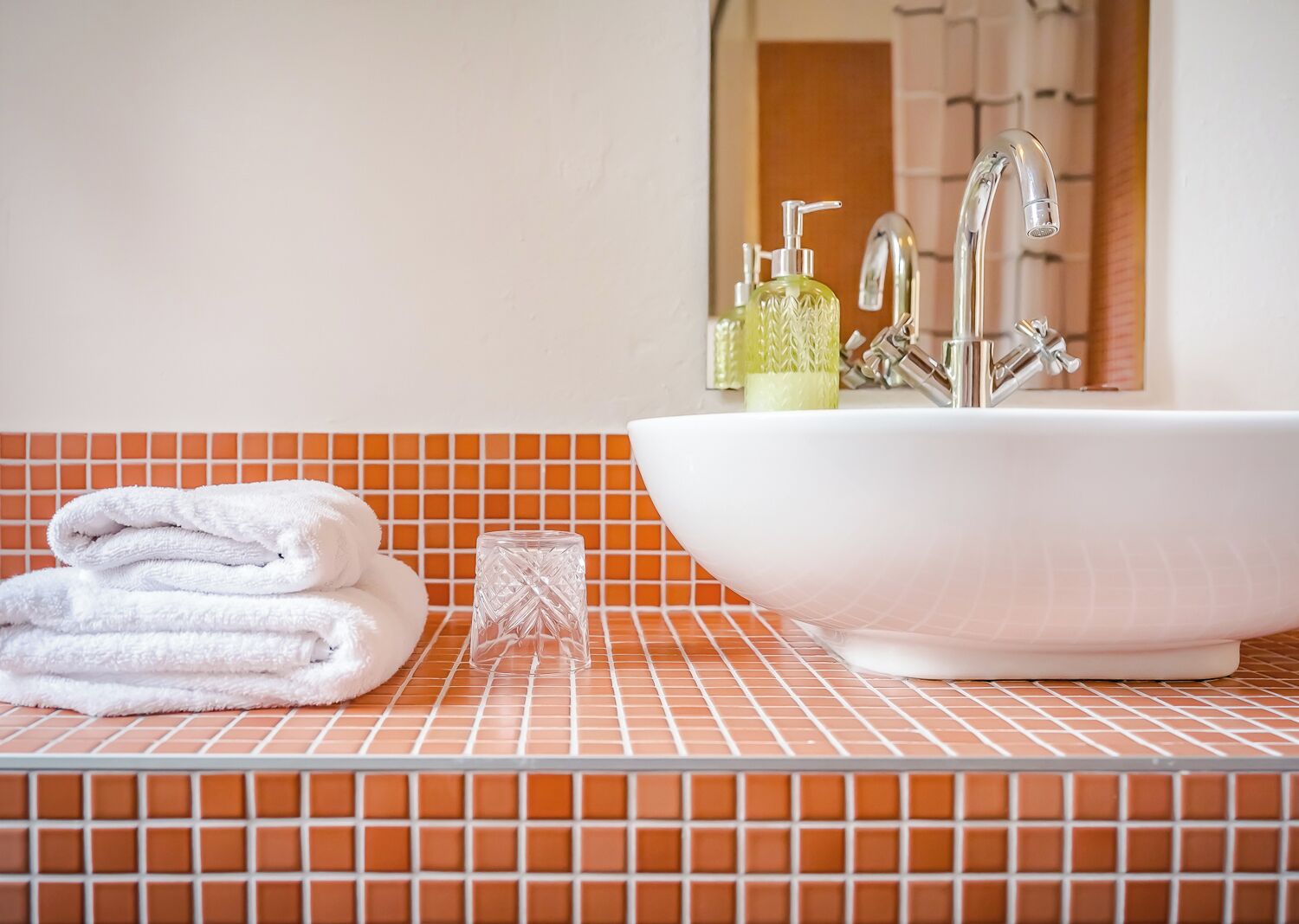 Casa de banho com lavatório branco, um copo de água e uma pilha de toalhas brancas sobre uma bancada em mosaico.