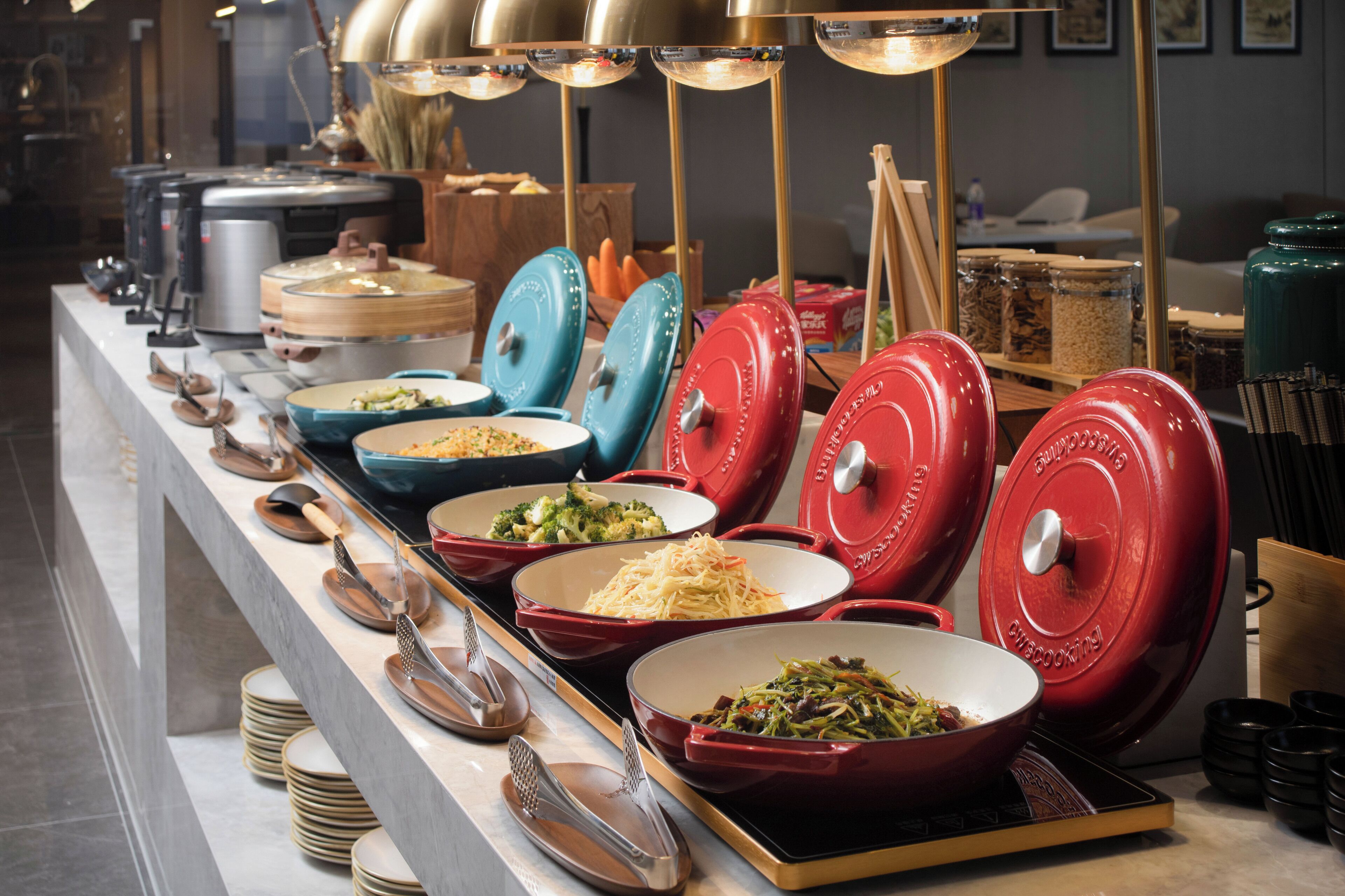 Disposição de buffet com diversos pratos de comida em recipientes vermelhos e azuis, dispostos sobre um balcão de mármore.