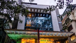 Entrada de um hotel com uma escadaria que leva à porta, uma placa com o nome do hotel e uma grande janela de vidro que reflete o ambiente.