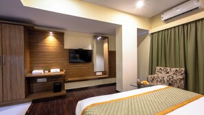 Quarto de hotel com cama, televisão, escrivaninha com uma planta e uma cadeira.