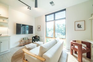 Uma sala de estar moderna com um sofá branco, uma cadeira de madeira, uma TV de tela plana na parede e janelas amplas com vista para o exterior.