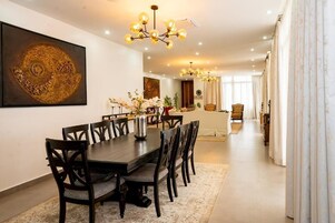 Uma sala de jantar com uma mesa comprida, cadeiras e um candelabro. Há um quadro grande na parede e uma janela com cortinas.