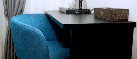 Canto de um quarto com uma cadeira azul, uma mesa preta, uma cafeteira e uma caixa decorativa.