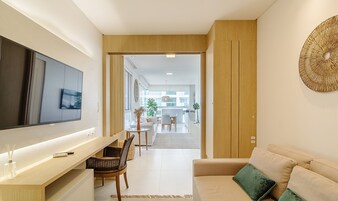 Uma sala moderna com televisão de tela plana, um sofá, uma área de jantar com mesa e cadeiras, e uma cozinha visível através de uma porta aberta.