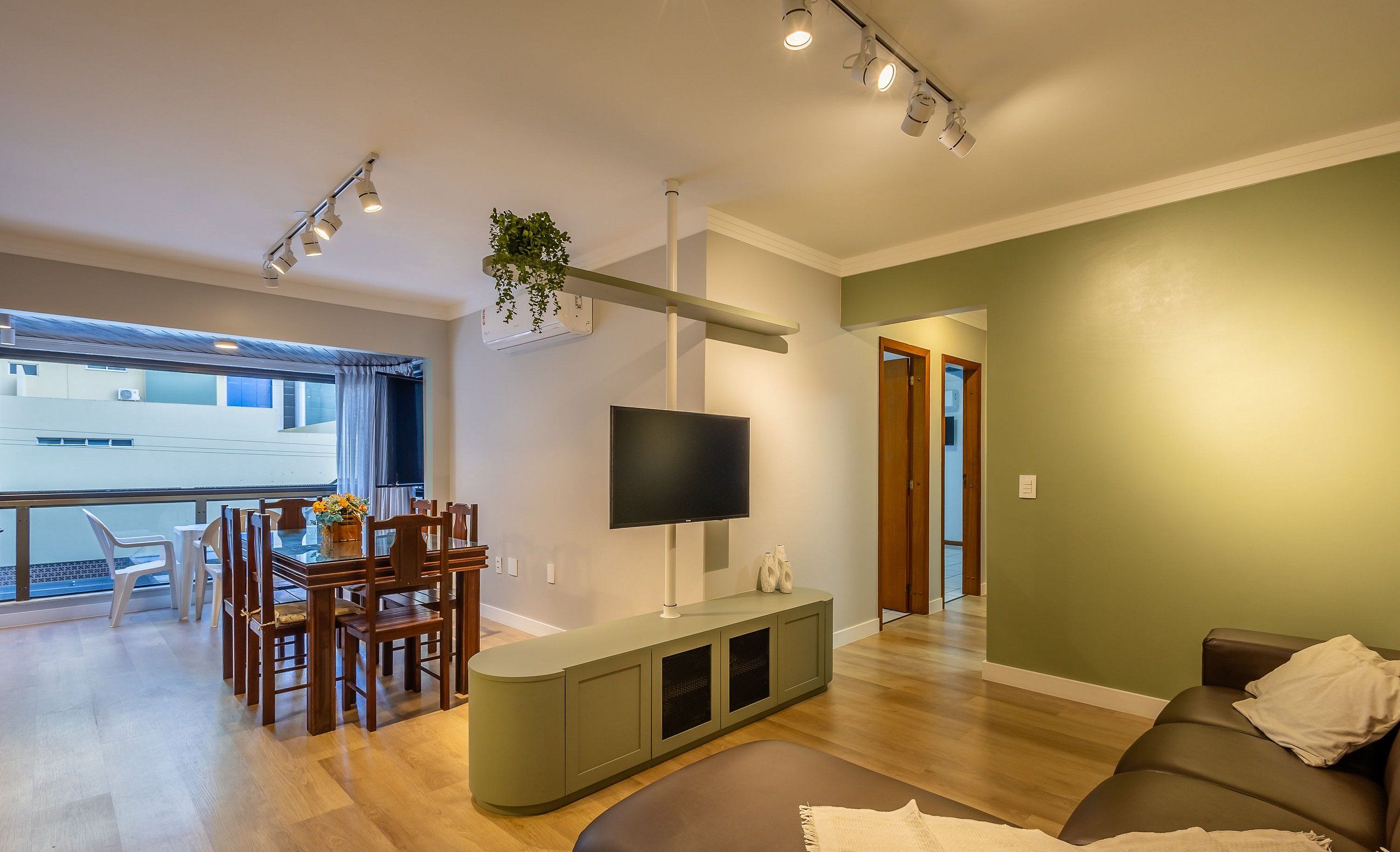 Uma sala de estar moderna com área de jantar, uma televisão de tela plana na parede e uma janela grande que oferece vista para o exterior.