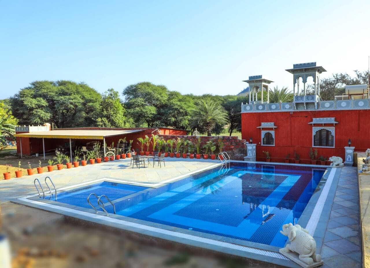 Uma piscina retangular com deck revestido, rodeada por plantas em vasos e um prédio vermelho com detalhes arquitetônicos.