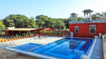 Uma piscina retangular com deck revestido, rodeada por plantas em vasos e um prédio vermelho com detalhes arquitetônicos.