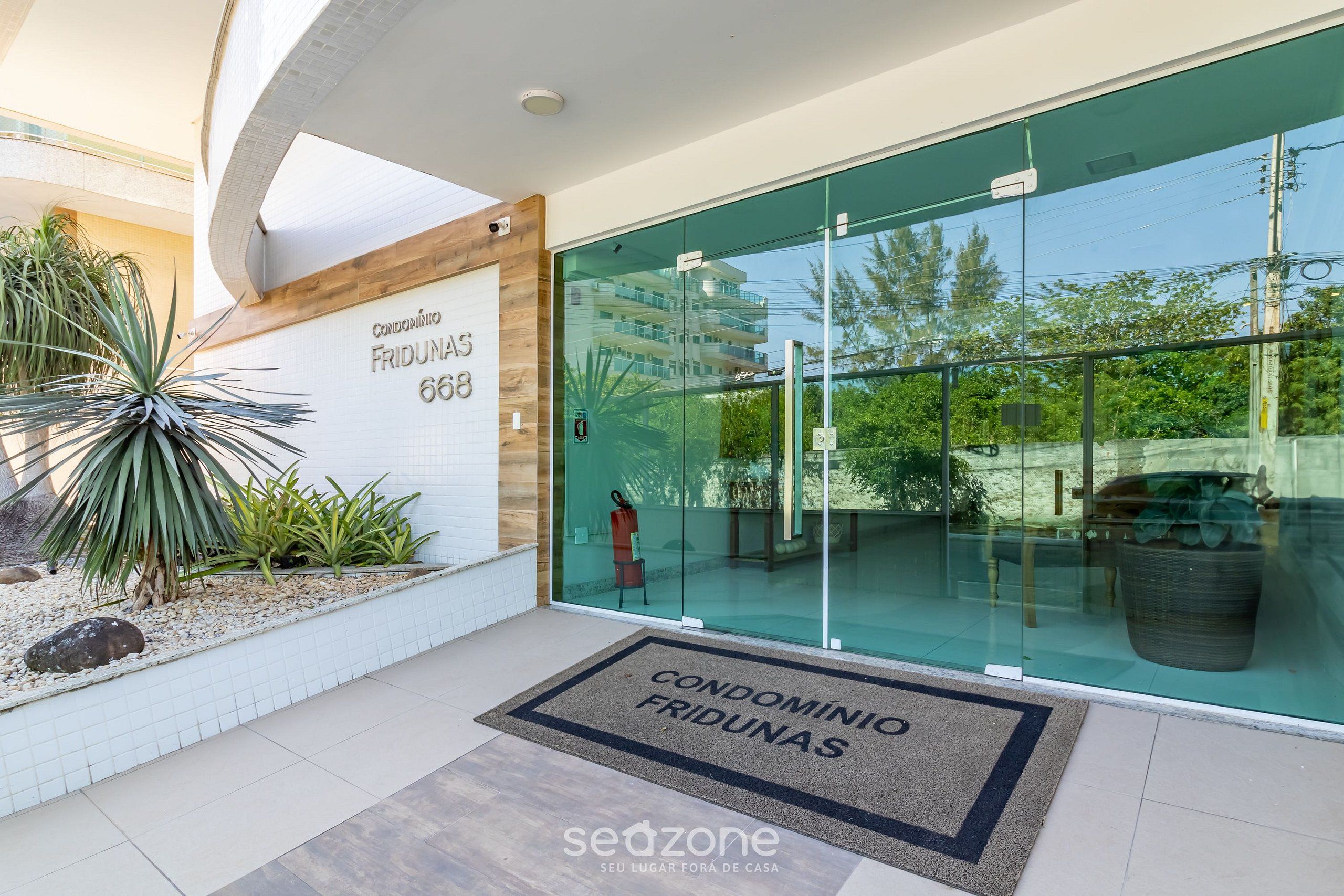 Entrada de um edifício moderno com porta de vidro, tapete de boas-vindas com o nome do edifício e um pequeno jardim com plantas.