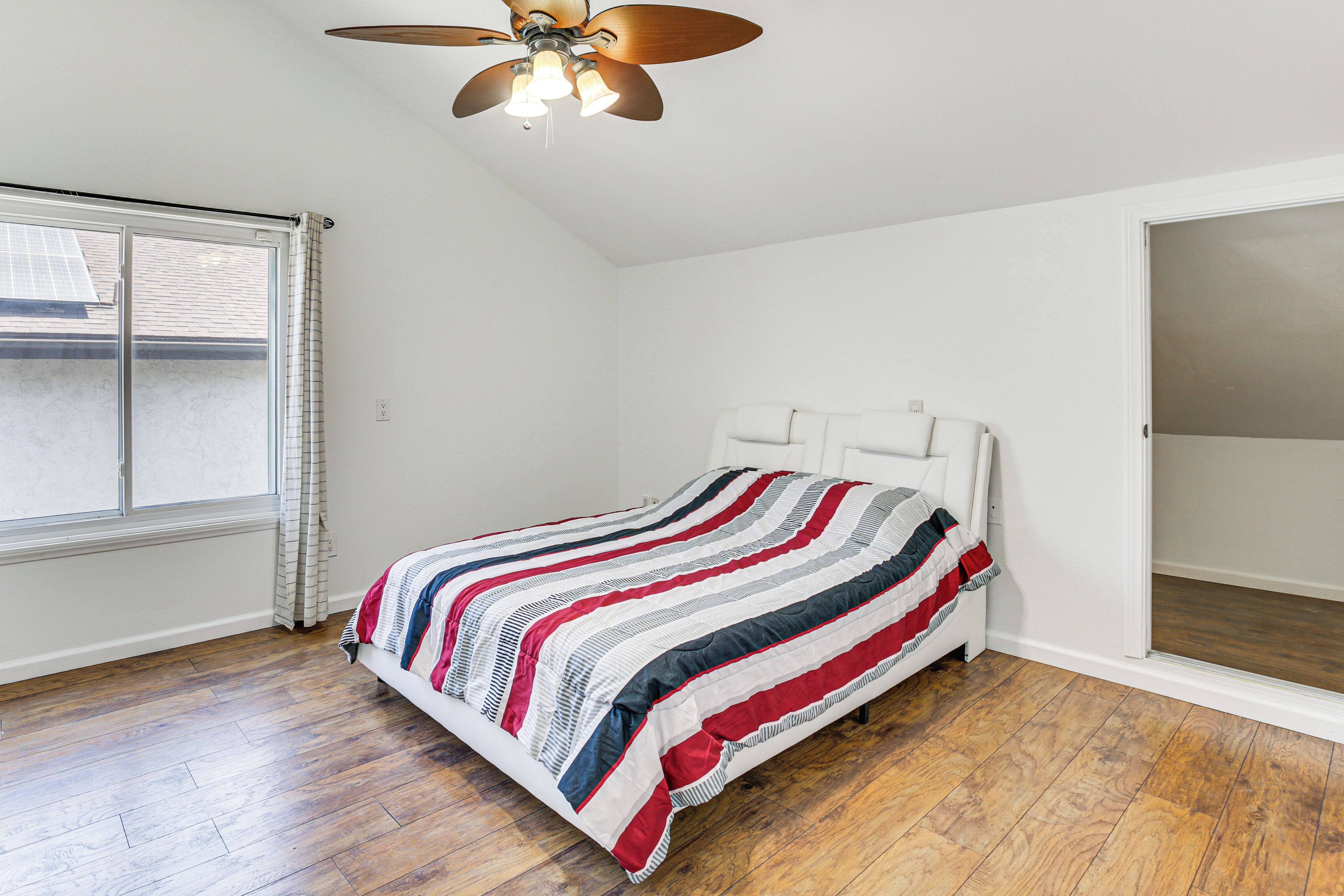 Quarto com cama, piso de madeira, ventilador de teto e janela com cortinas.