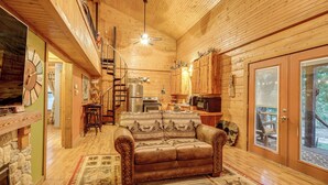 Interior acolhedor de uma cabana, com área de estar central que inclui um sofá de couro, pisos de madeira, lareira e escada para um andar superior.