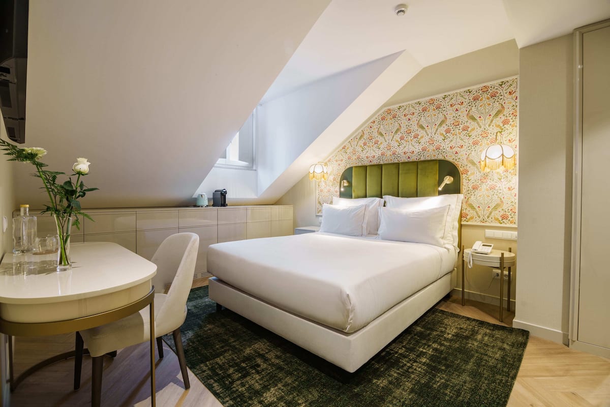 Quarto moderno com uma cama grande, uma mesinha redonda, uma cadeira, um criado-mudo com abajur e um detalhe decorativo na parede.