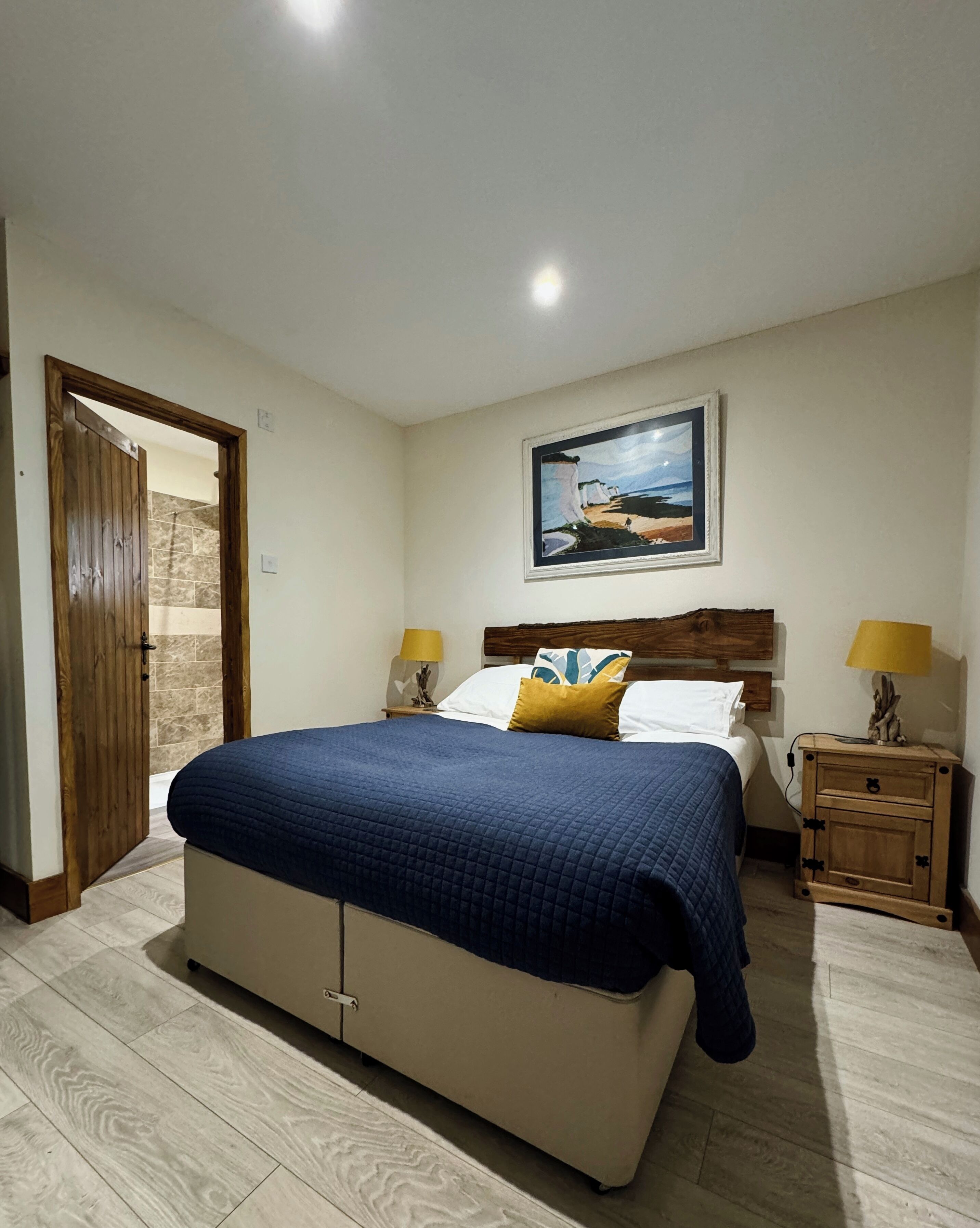Quarto com cama, cabeceira de madeira, duas mesas de cabeceira com luminárias, um quadro na parede e uma porta que dá para outro cômodo.
