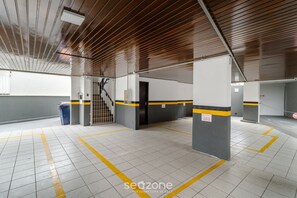 Garagem de estacionamento com vagas demarcadas, escada e um contentor de lixo.