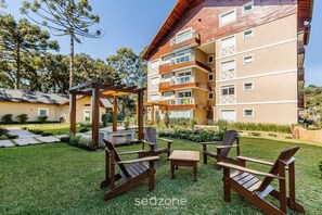 Uma área externa com assentos de madeira, cadeiras e uma mesa, rodeada por um gramado bem cuidado e um edifício com várias varandas.