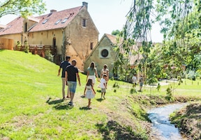 Uma família caminhando por um caminho gramado ao lado de um riacho, com um edifício de pedra e árvores ao fundo.